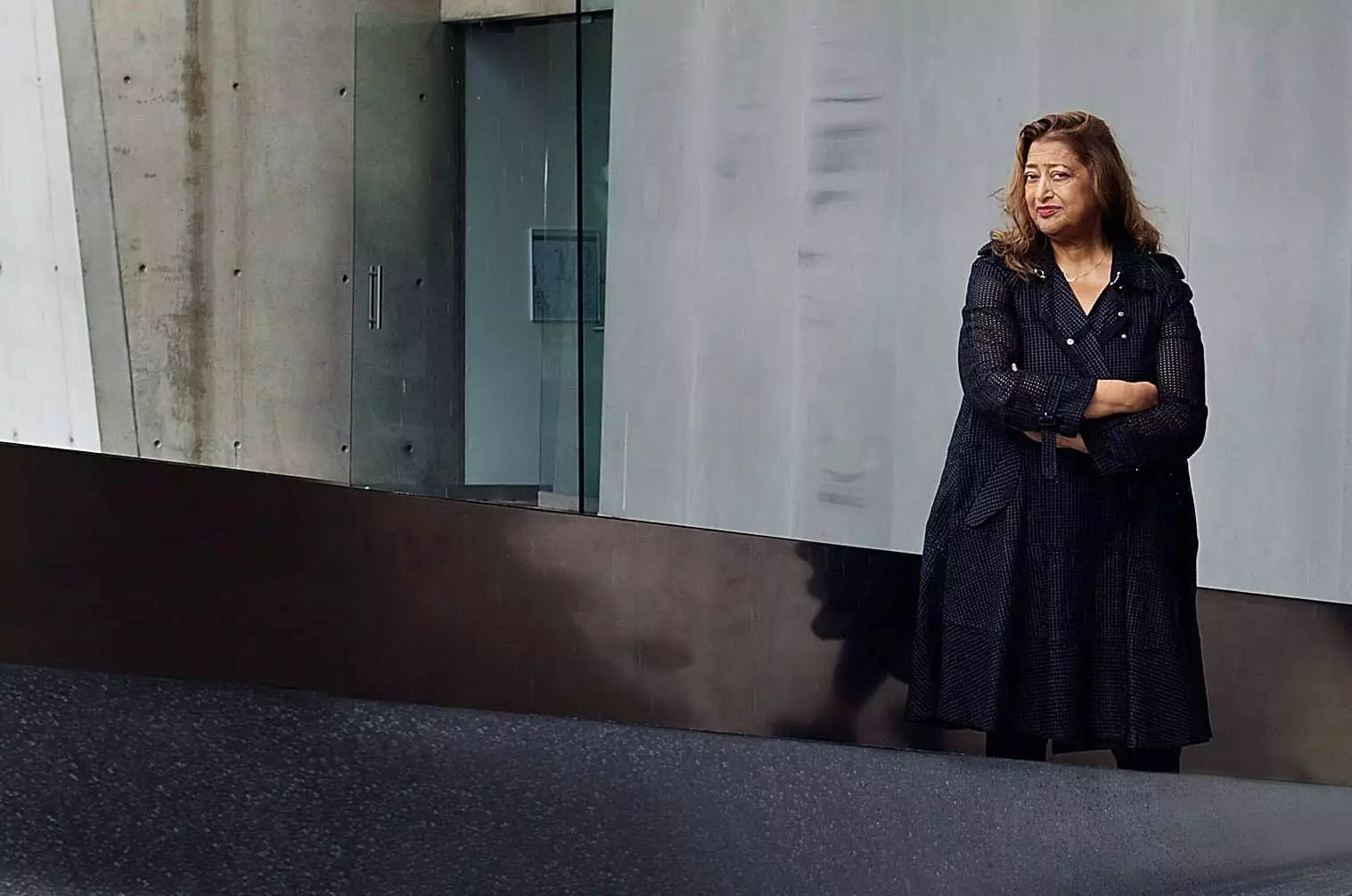 argitek Zaha Hadid, lang donker hare, arms gevou, stanind voor grys gebou en blink beeldhouwerk