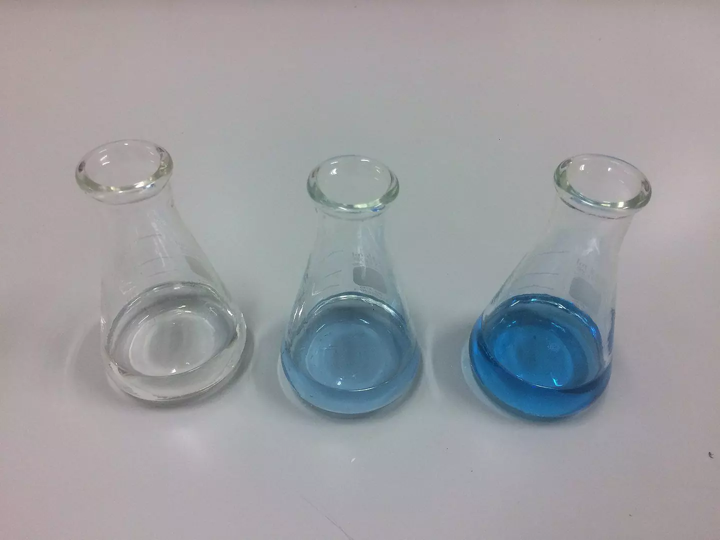 Tiga gelas kimia berbeda dengan cairan di dalamnya dengan latar belakang abu-abu.