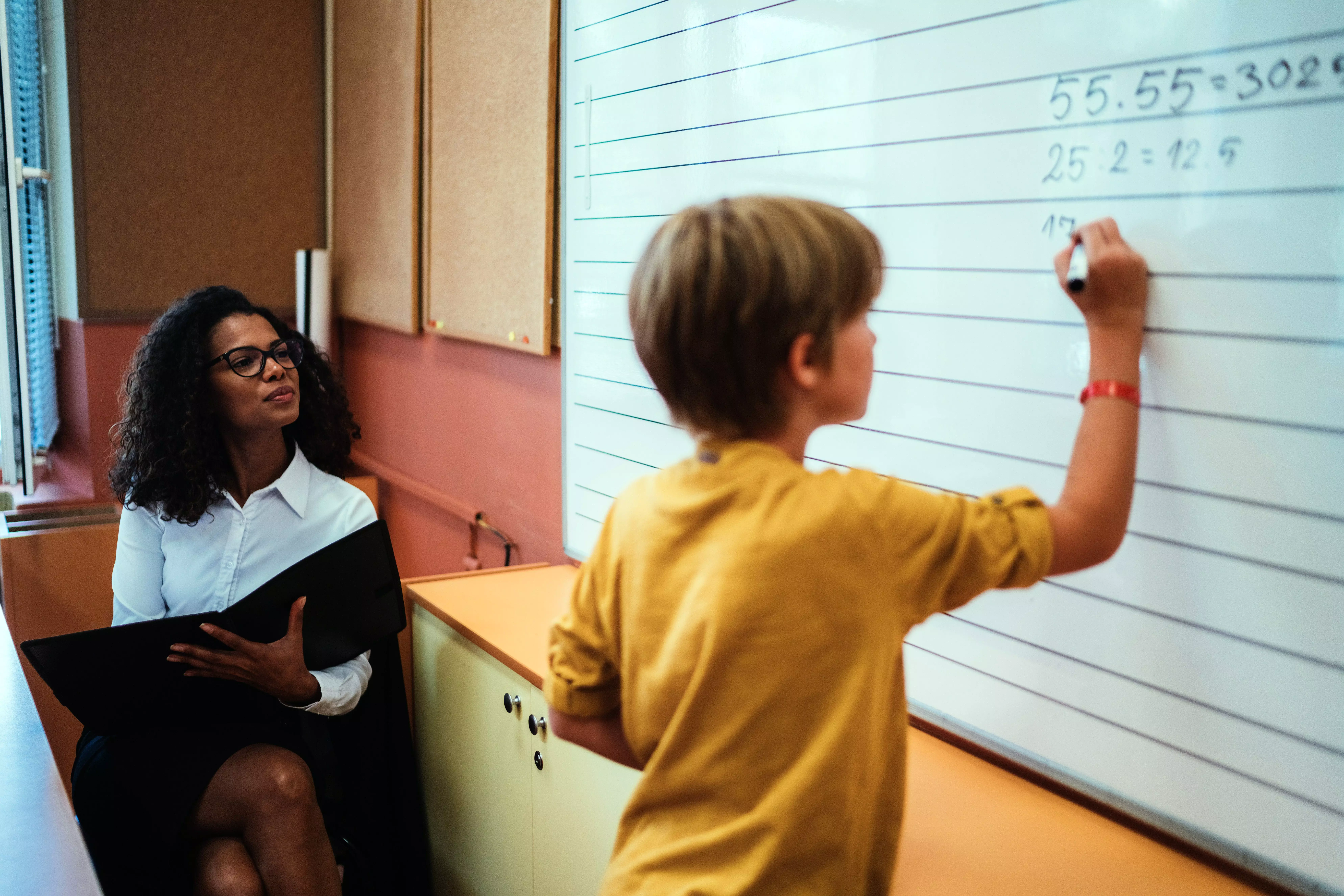 Un băiat face probleme de matematică pe tablă sub privirea atentă a profesorului