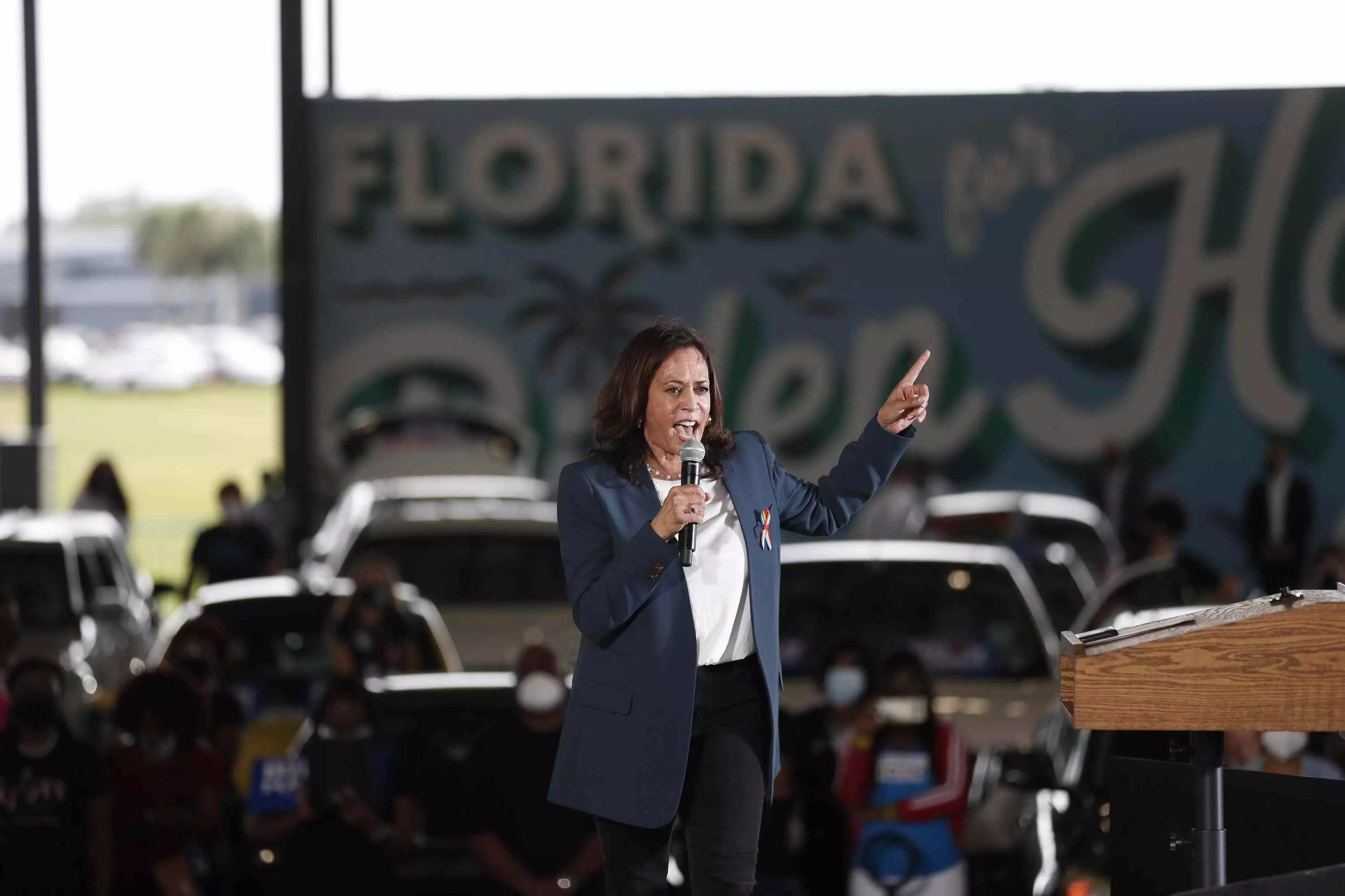 Kamala Harris vorbind la microfon și arătând cu degetul în timp ce stă pe o scenă înconjurată de mașini și de membri ai publicului din Florida