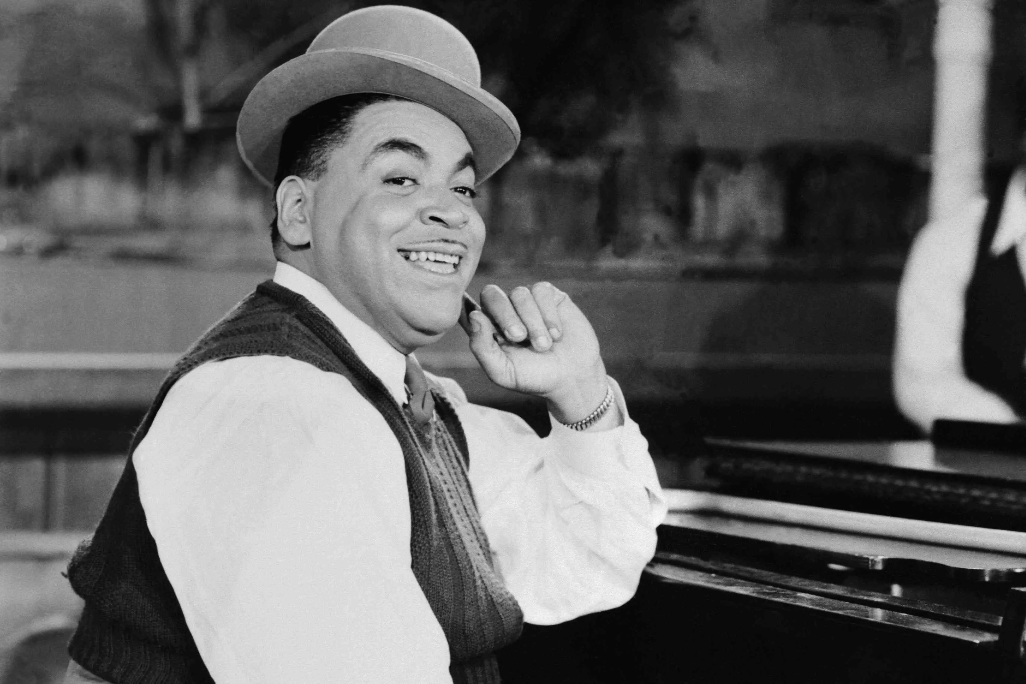 Fats Waller se naslanja na klavir in se smehlja, nosi klobuk in telovnik