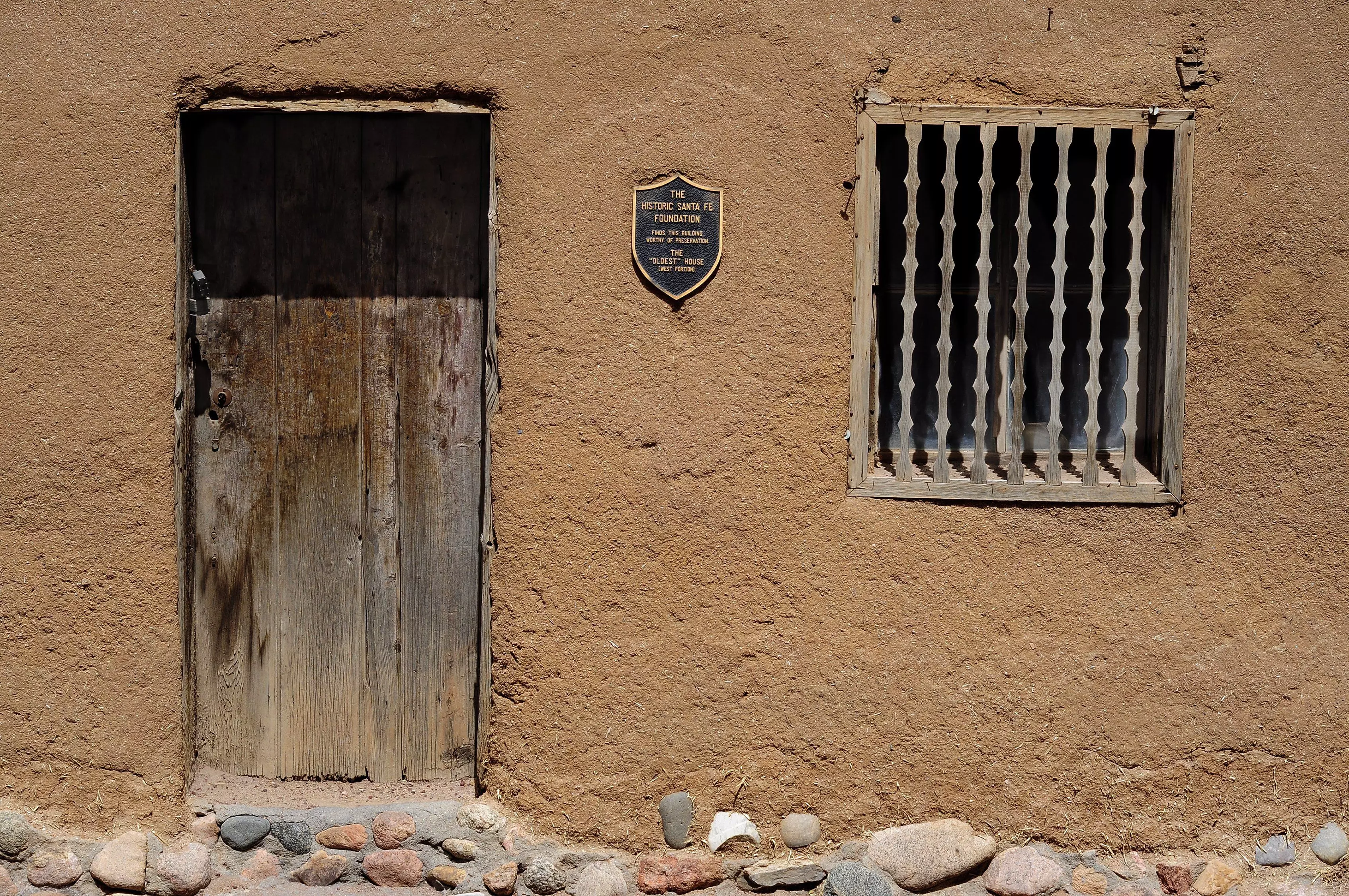 Nahaufnahmedetail einer hellbraunen Adobe-Fassade mit Holztür, vertikalen Balken an einem Fenster und einer schildförmigen Plakette dazwischen