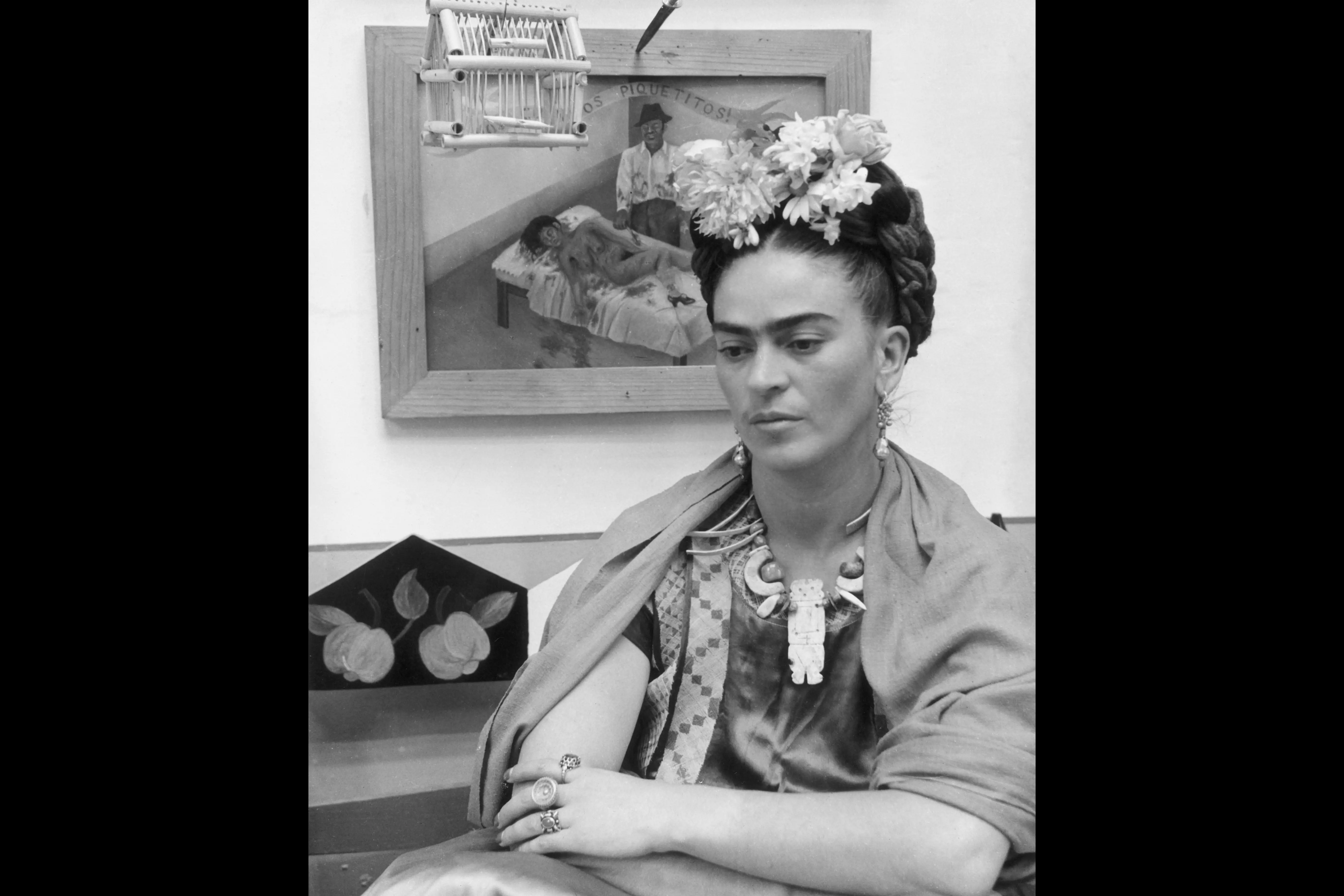 Den meksikanske maleren Frida Kahlo sitter med armene foldet og ser ned foran et av maleriene hennes og et fuglebur i tre.