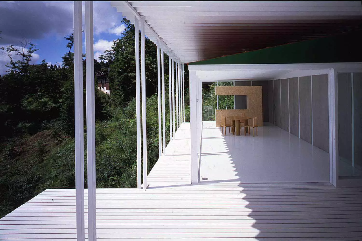 Blick hinunter zum Ende eines kahlen, weißen Raums, zwei Decken, offene Wand links mit Pfosten, Blick auf den Wald