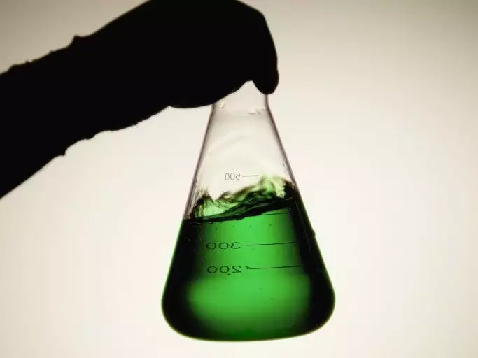 Una mano enguantada hace girar un matraz Erlenmeyer que contiene un líquido verde.