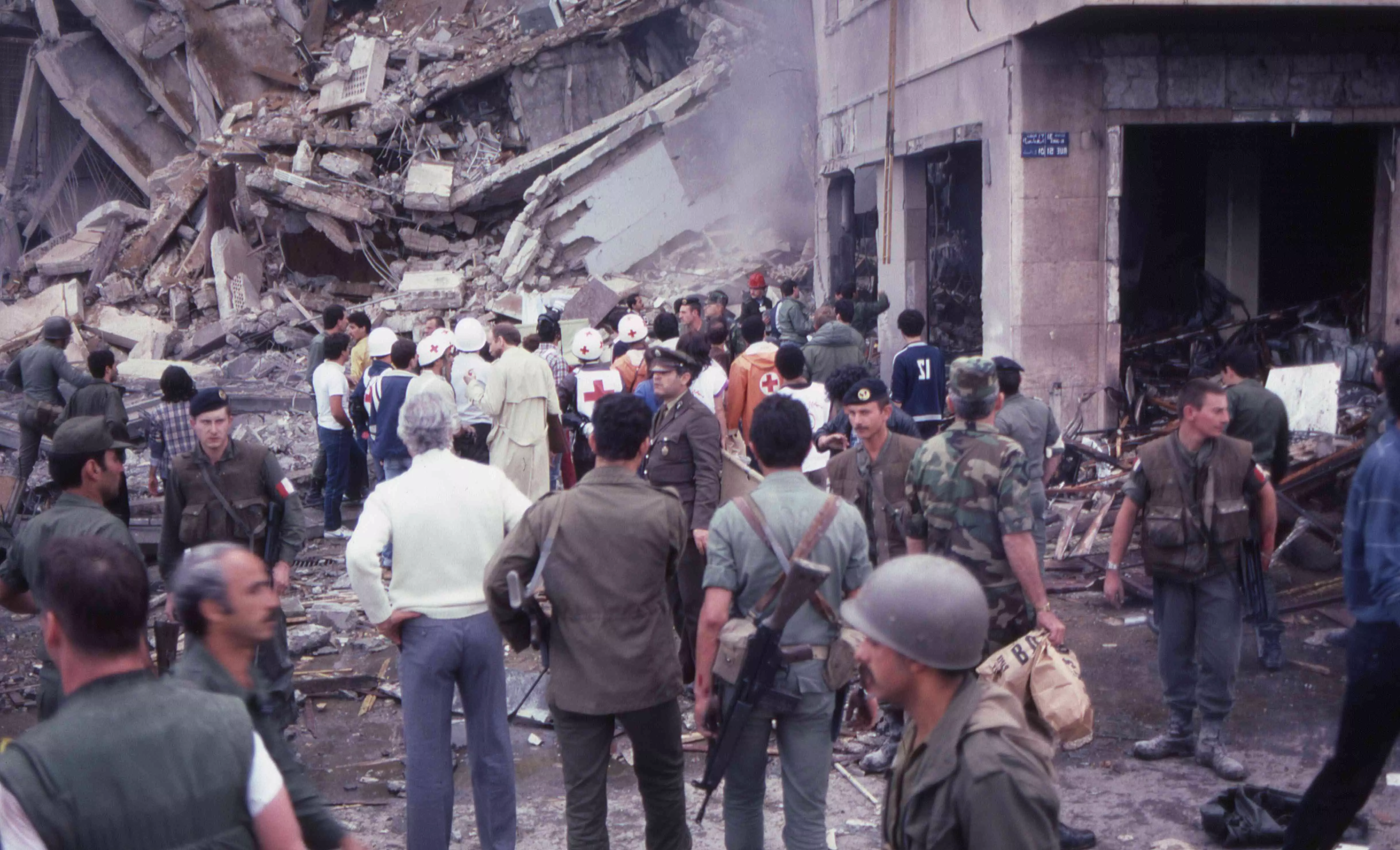 Тълпа войници и помощници стоят сред разрушенията и щетите на мястото на самоубийствения атентат в американското посолство, Бейрут, Ливан, 18 април 1983 г.