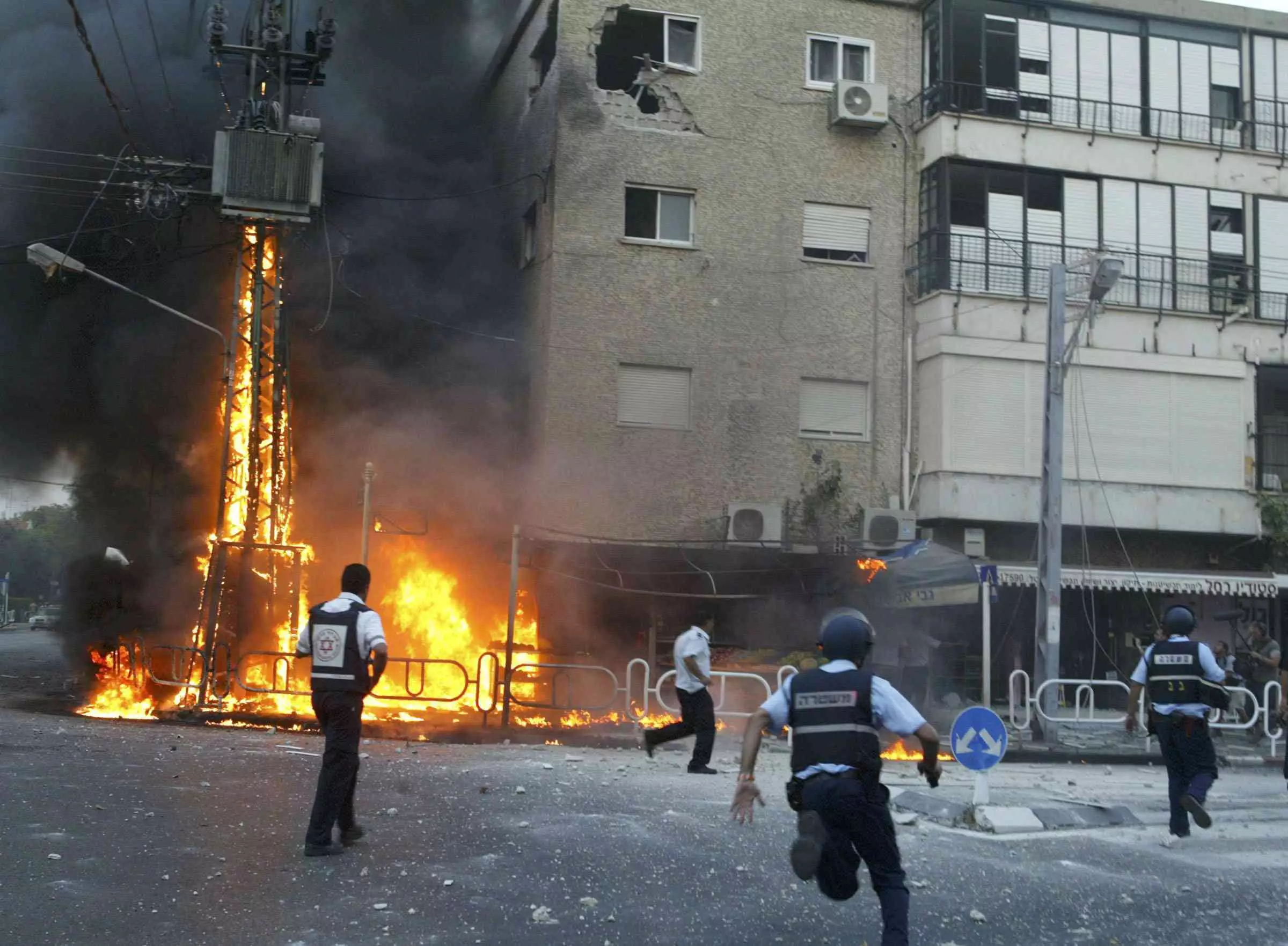 Израелски полицаи се втурват към мястото на горящ електрически стълб и повредена сграда моменти след залп от ракети на Хизбула на 13 юли 2006 г. в северния град Нахария, Израел.