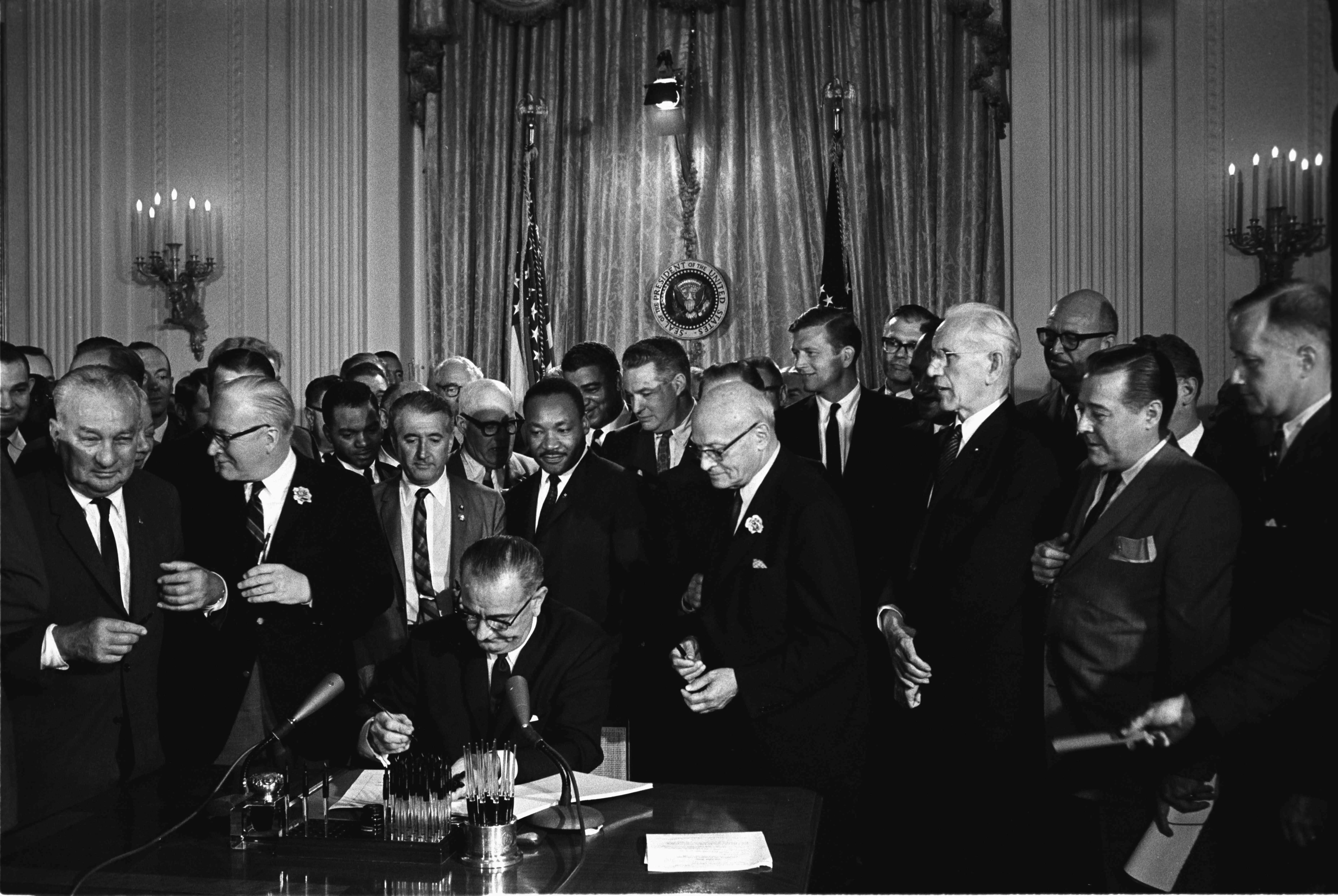 O presidente Lyndon B. Johnson asina a Lei de Dereitos Civís de 1964 mentres Martin Luther King, Jr. e outros observan.