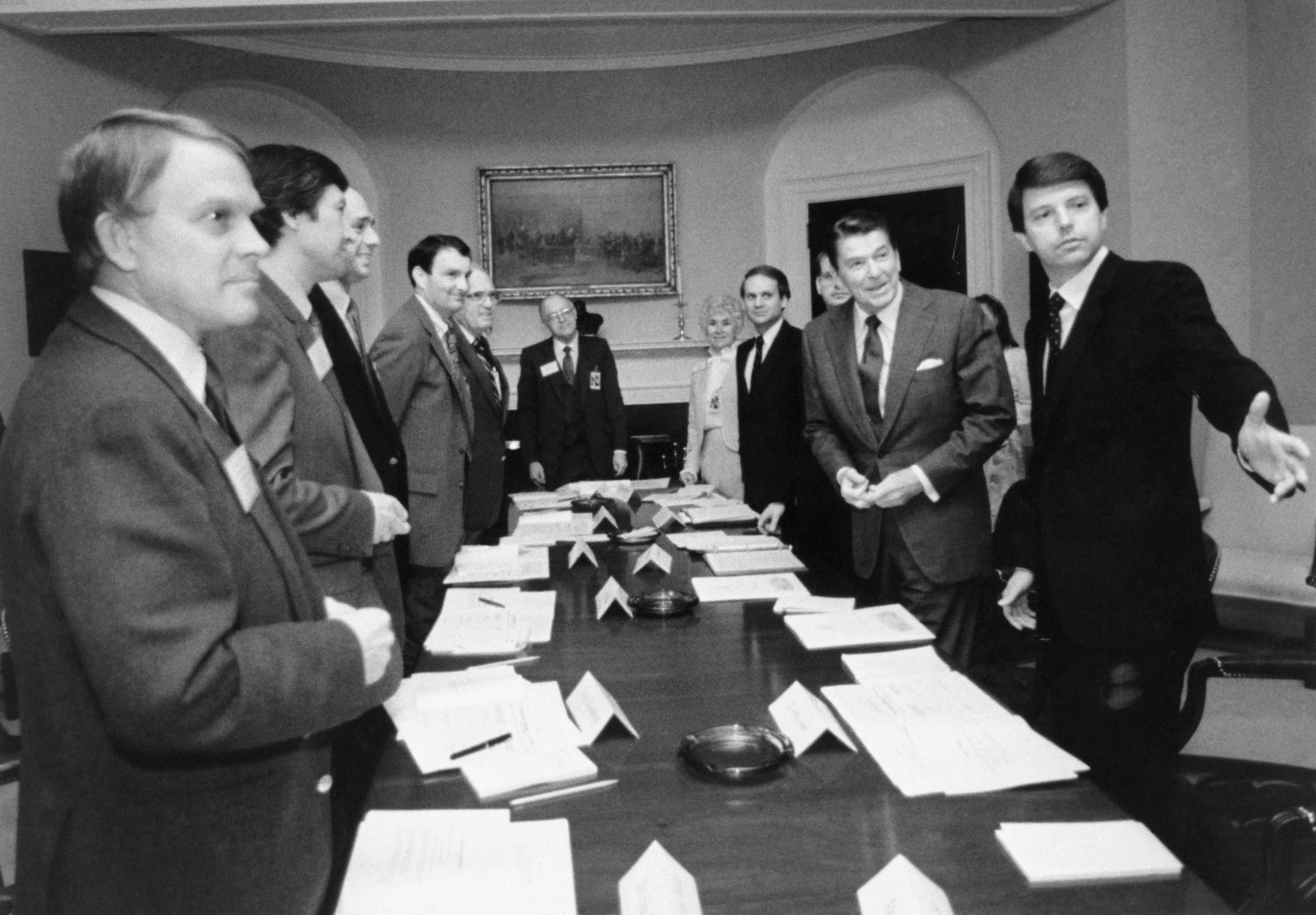 Nigra kaj blanka bildo de Ronald Reagan kaj pluraj aliaj viroj en vestokompletoj ĉirkaŭ longa konferenca tablo