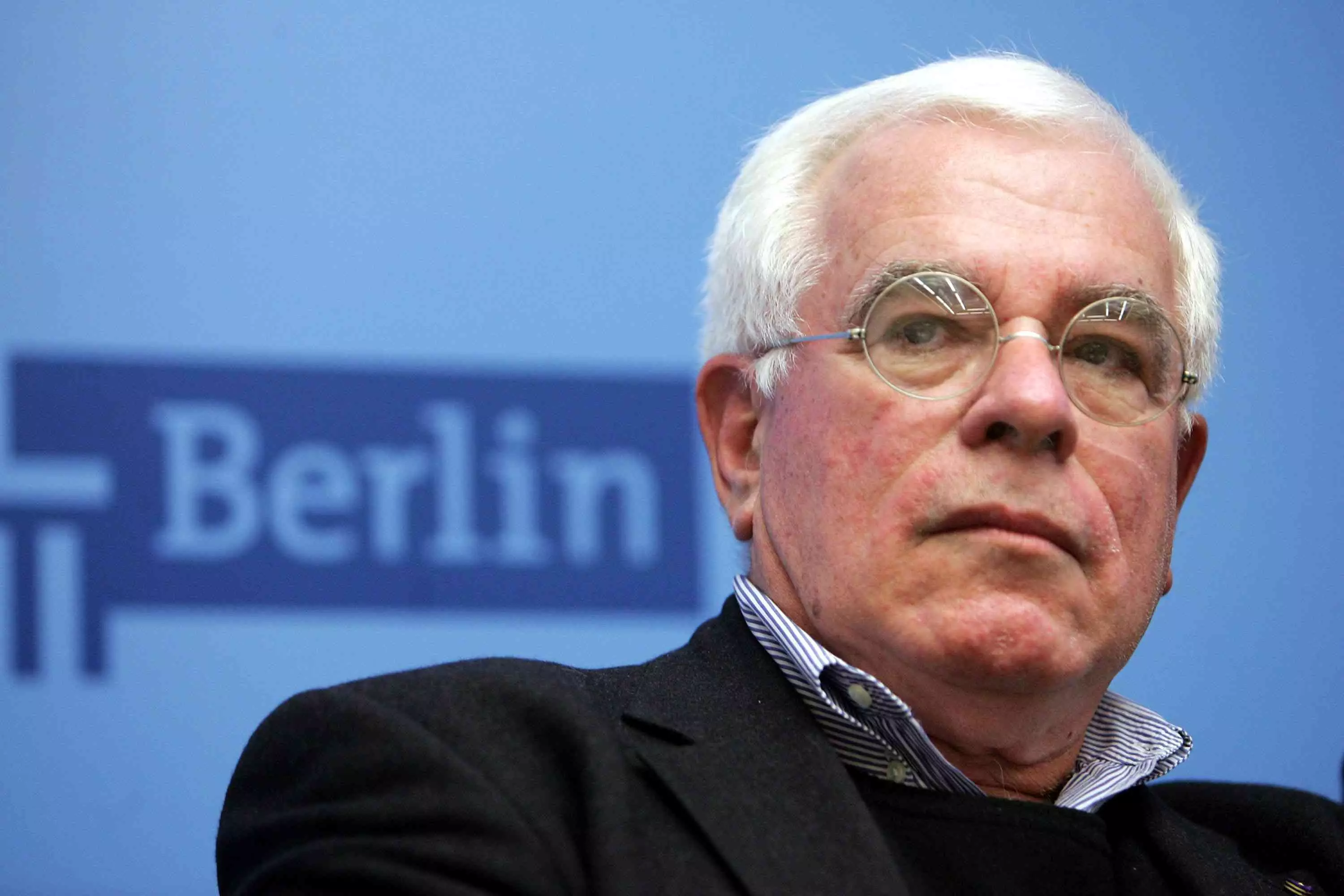 weißer Mann, weißes Haar, dünnrandige Brille, Berlin-Schild im Hintergrund